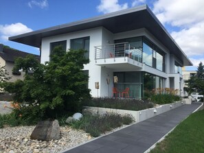 Uma casa moderna de dois andares com varanda, jardim com pedras e plantas, e um caminho pavimentado.