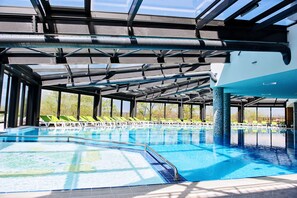 Piscina com água azul cristalina, rodeada por espreguiçadeiras verdes e uma área coberta com vigas de metal e painéis de vidro.
