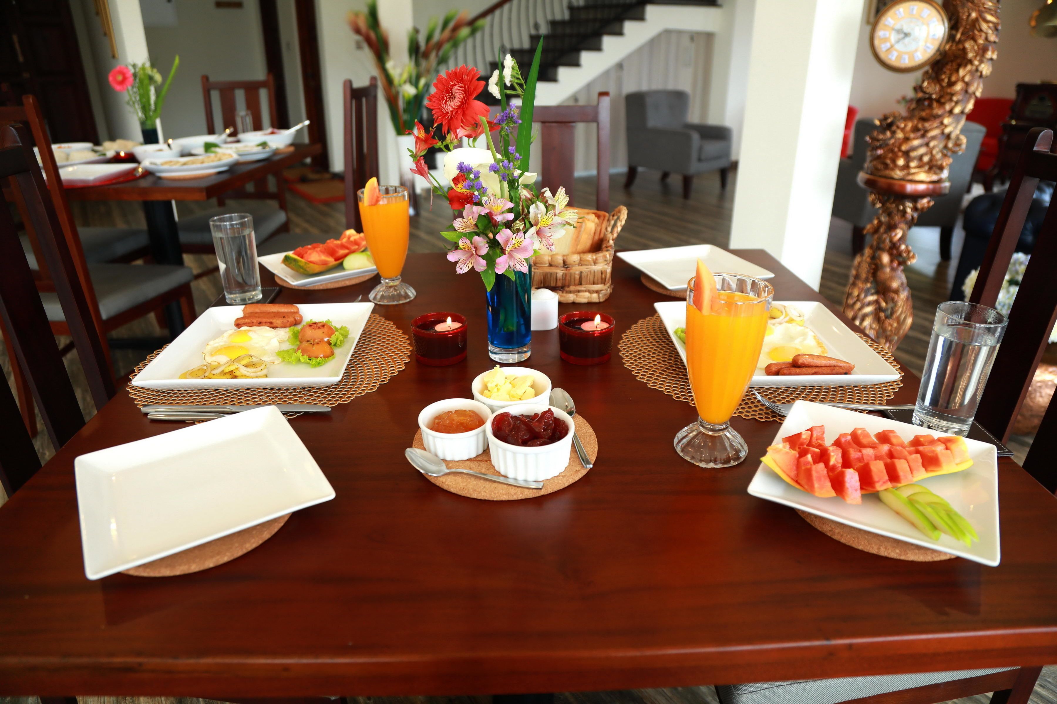 Mesa de jantar posta com itens de café da manhã, incluindo pratos com ovos, torradas e frutas, além de um vaso de flores e velas.
