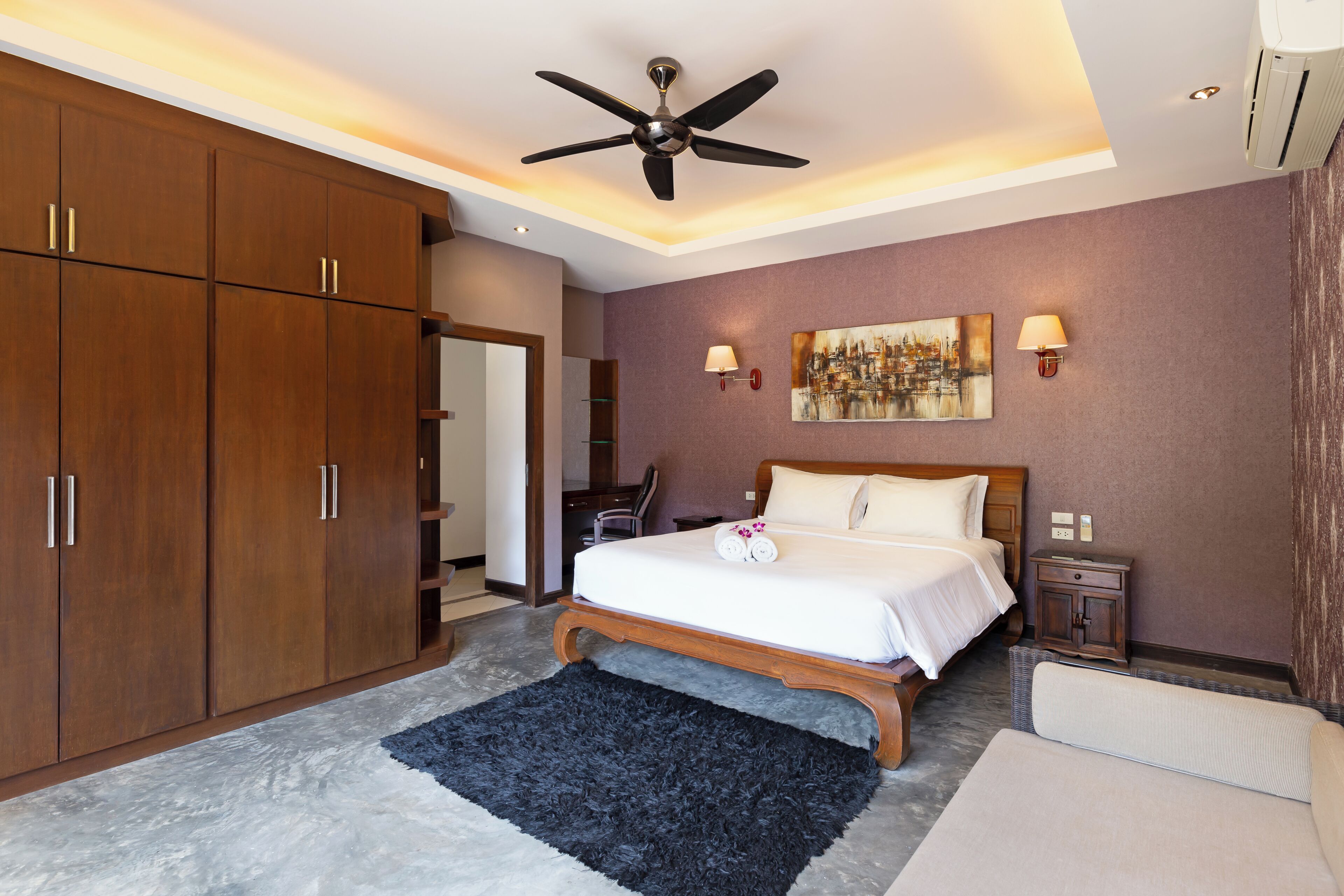 Quarto com cama, ventoinha no teto, mesa de cabeceira, escrivaninha e guarda-roupa.
