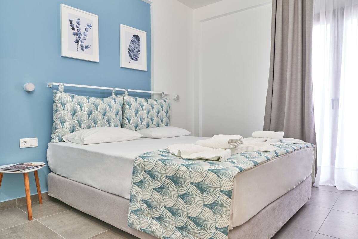 Uma cama bem arrumada com um cobertor estampado com padrão de folha, uma pequena mesa de cabeceira com uma revista e duas obras de arte emolduradas na parede.