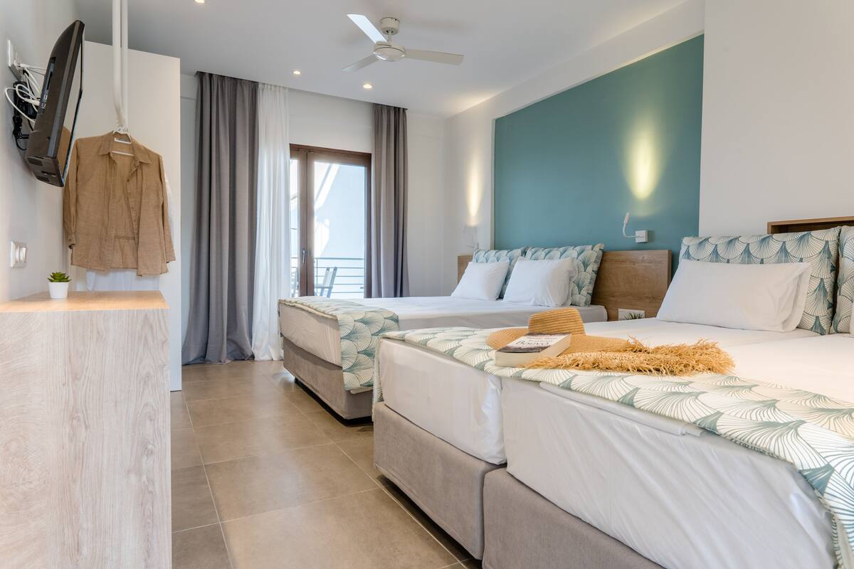 Quarto de hotel com duas camas, televisão fixada na parede e varanda com cortinas.