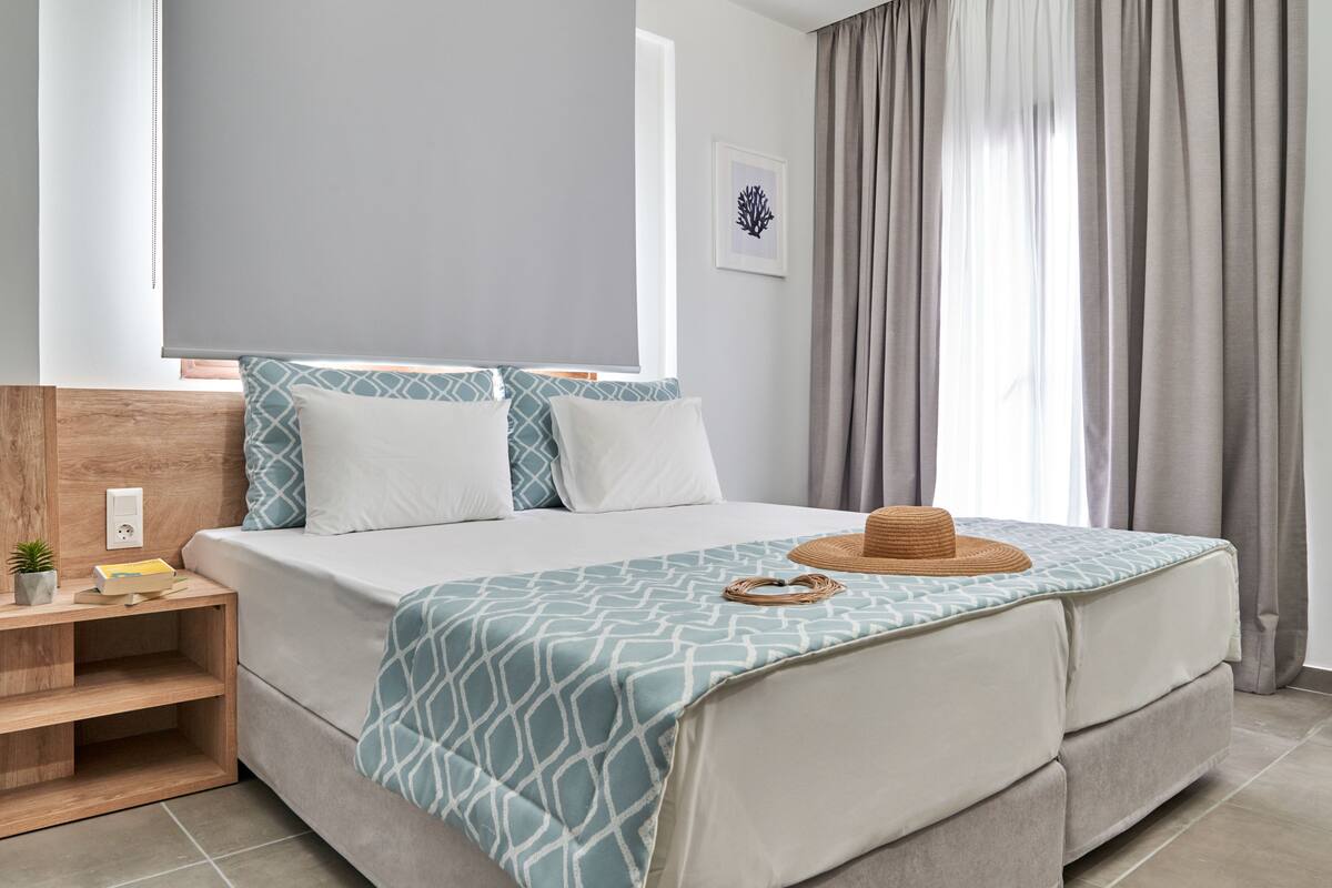 Uma cama bem arrumada com colcha estampada, uma mesa de cabeceira com abajur e uma janela com cortinas.