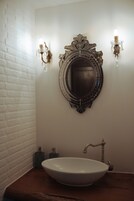 Casa de banho com parede de tijolos brancos, espelho redondo com detalhes ornamentais e duas luzes de parede.