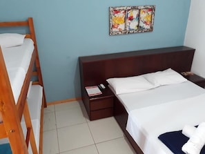 Quarto de hotel com uma cama beliche, uma cama de solteiro, uma mesa de cabeceira com telefone e um livro, e uma obra de arte colorida na parede.