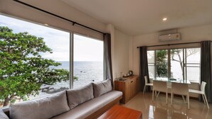 Quarto moderno com uma grande janela com vista para o mar, um sofá, uma mesa de jantar com cadeiras e uma pequena cozinha compacta.