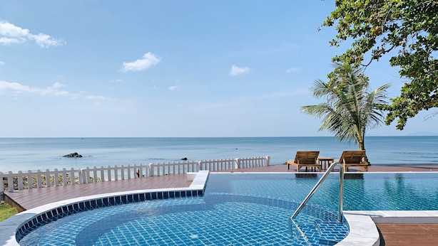 Piscina com deck de madeira e grade branca, com vista para uma praia com espreguiçadeiras e uma palmeira.