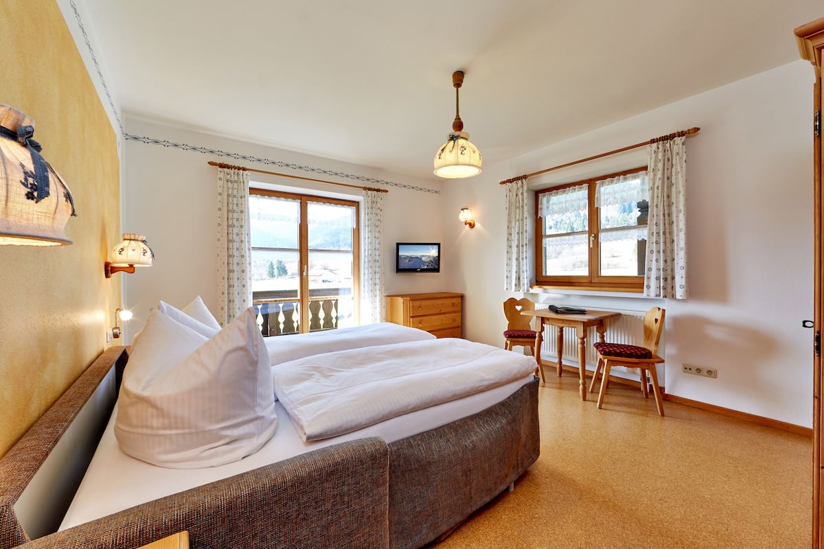 Quarto de hotel com uma cama grande, uma escrivaninha com cadeira, televisão e vista para o exterior pelas janelas.