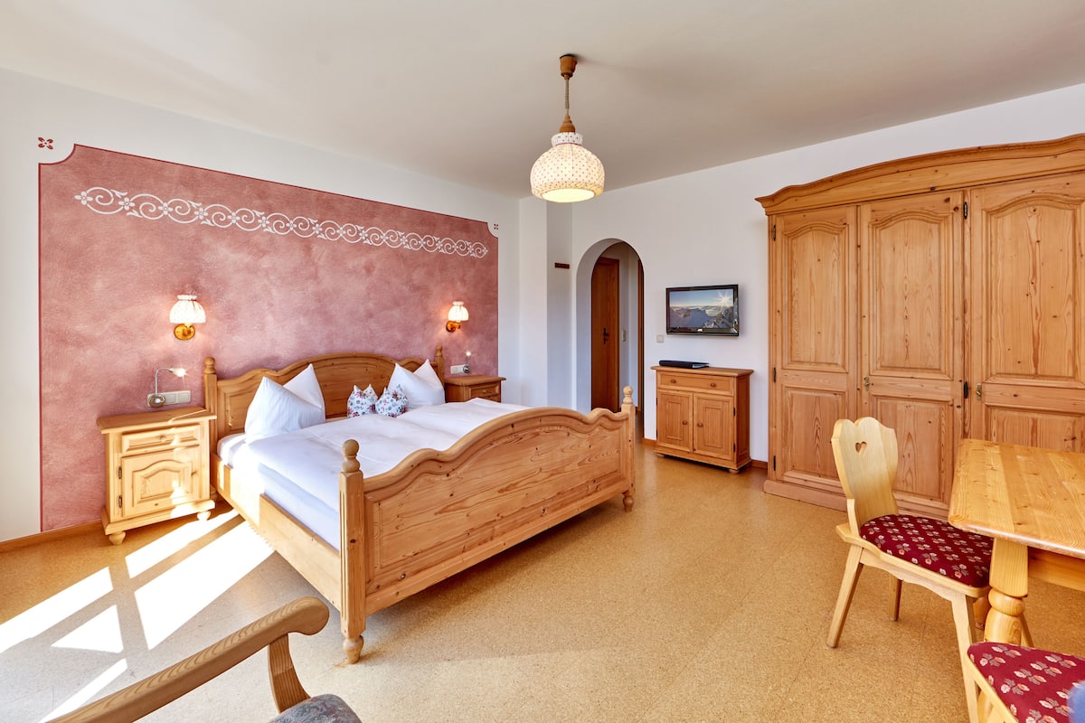 Quarto com cama de madeira, luminárias de cabeceira, guarda-roupa de madeira, escrivaninha de madeira com cadeira e televisão na parede.