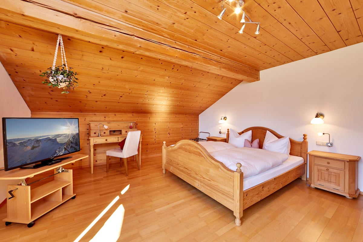 Quarto com teto de madeira, uma cama, televisão sobre um móvel, escrivaninha e mesa de cabeceira.