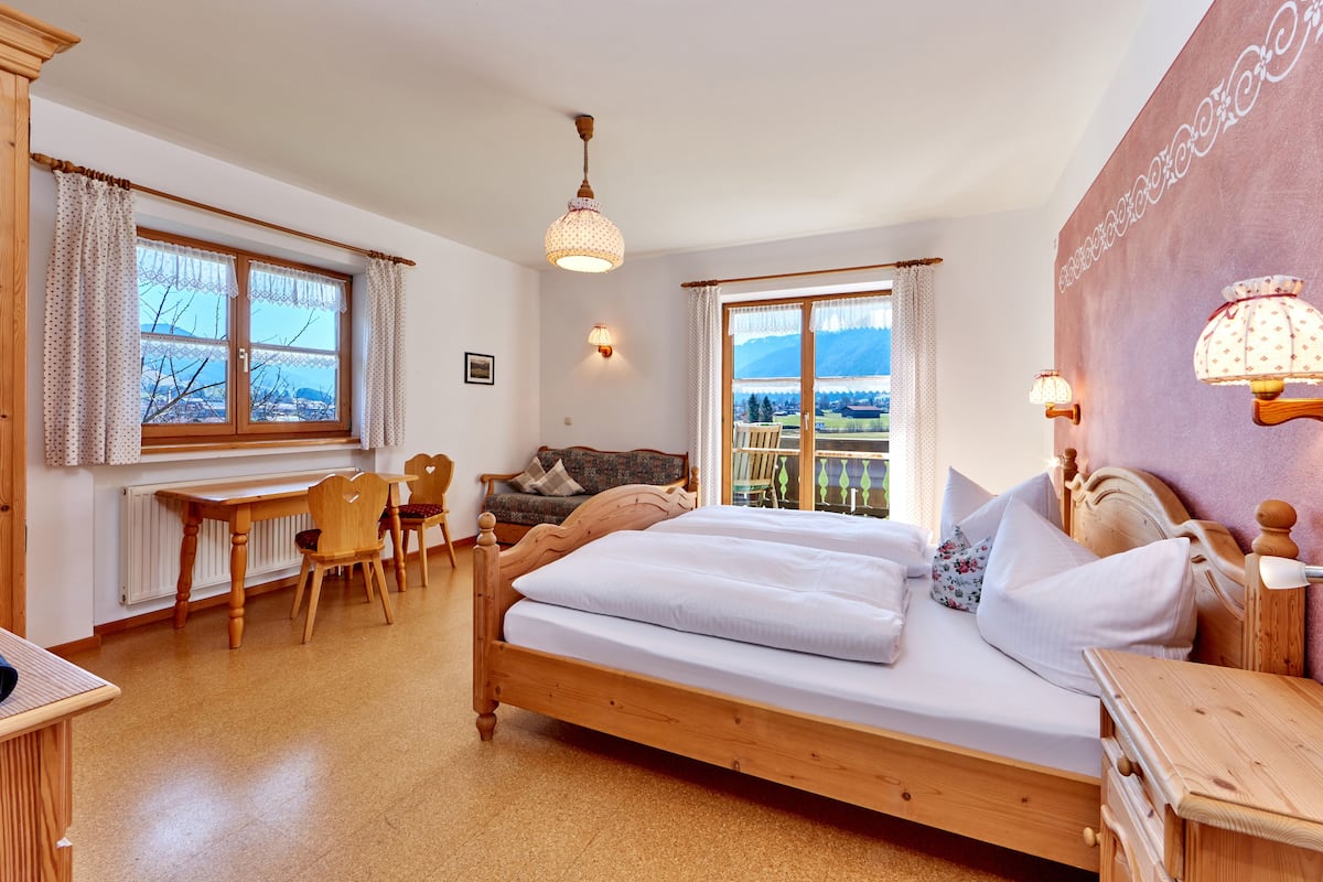 Quarto com cama de madeira, escrivaninha e cadeira, vista para o exterior através de grandes janelas e luminária fixa na parede.