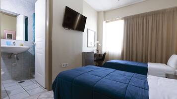 Quarto de hotel com cama, escrivaninha, cadeira, televisão, janela com cortinas e banheiro com pia e espelho.