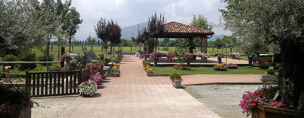 Um caminho de jardim leva a um gazebo com telhado de azulejos, cercado por canteiros de flores e árvores.