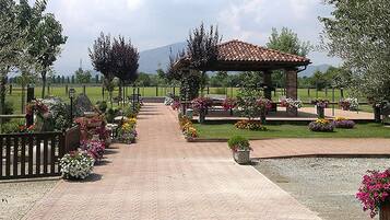 Um caminho de jardim leva a um gazebo com telhado de azulejos, cercado por canteiros de flores e árvores.