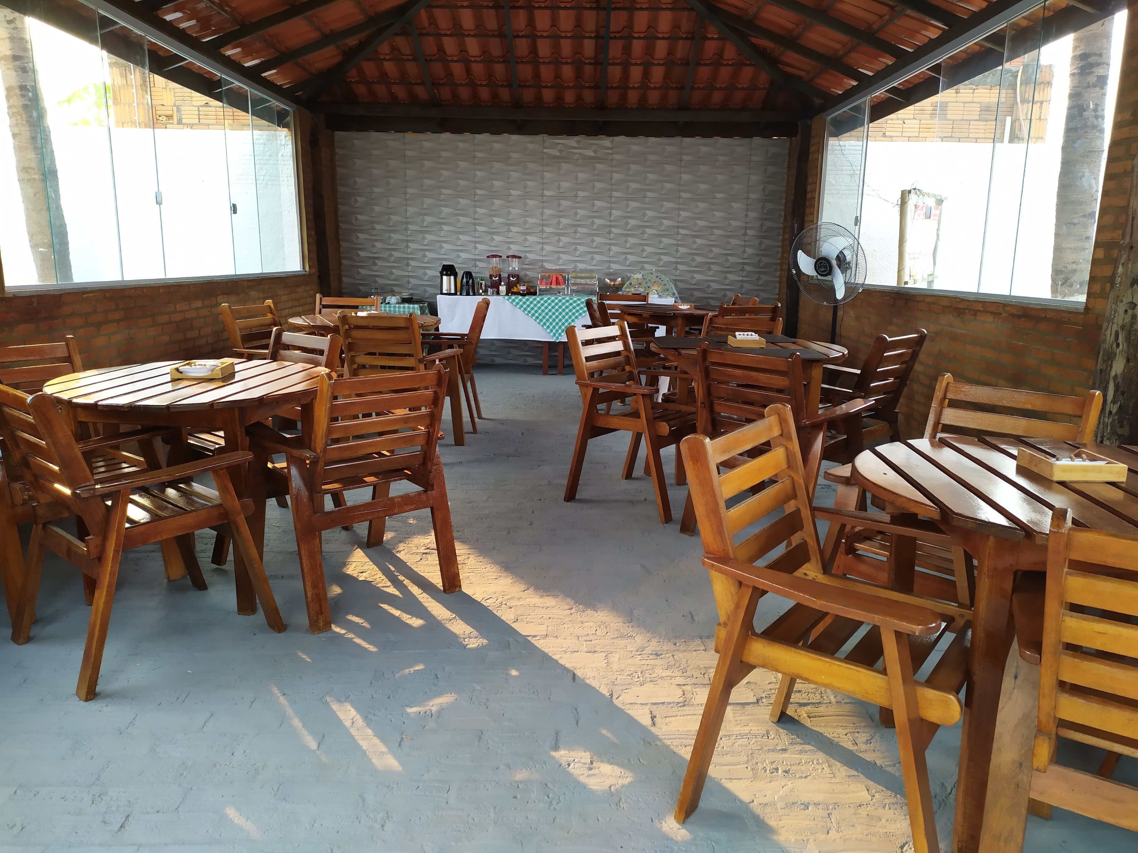 Uma área de refeições ao ar livre coberta, com mesas e cadeiras de madeira, uma parede de tijolos e um ventilador.