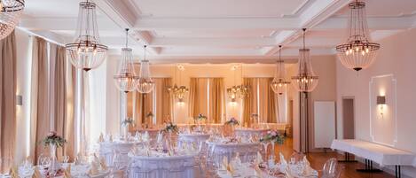 Uma sala de banquetes com mesas redondas, cadeiras transparentes e lustres de cristal.