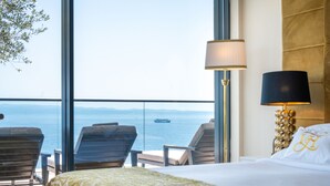 Quarto de hotel com uma grande janela que oferece vista para o mar e um navio. Há duas poltronas confortáveis e uma cama com colcha estampada.