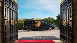 Uma van preta estacionada em frente a uma entrada com portão, com um tapete vermelho levando à porta.