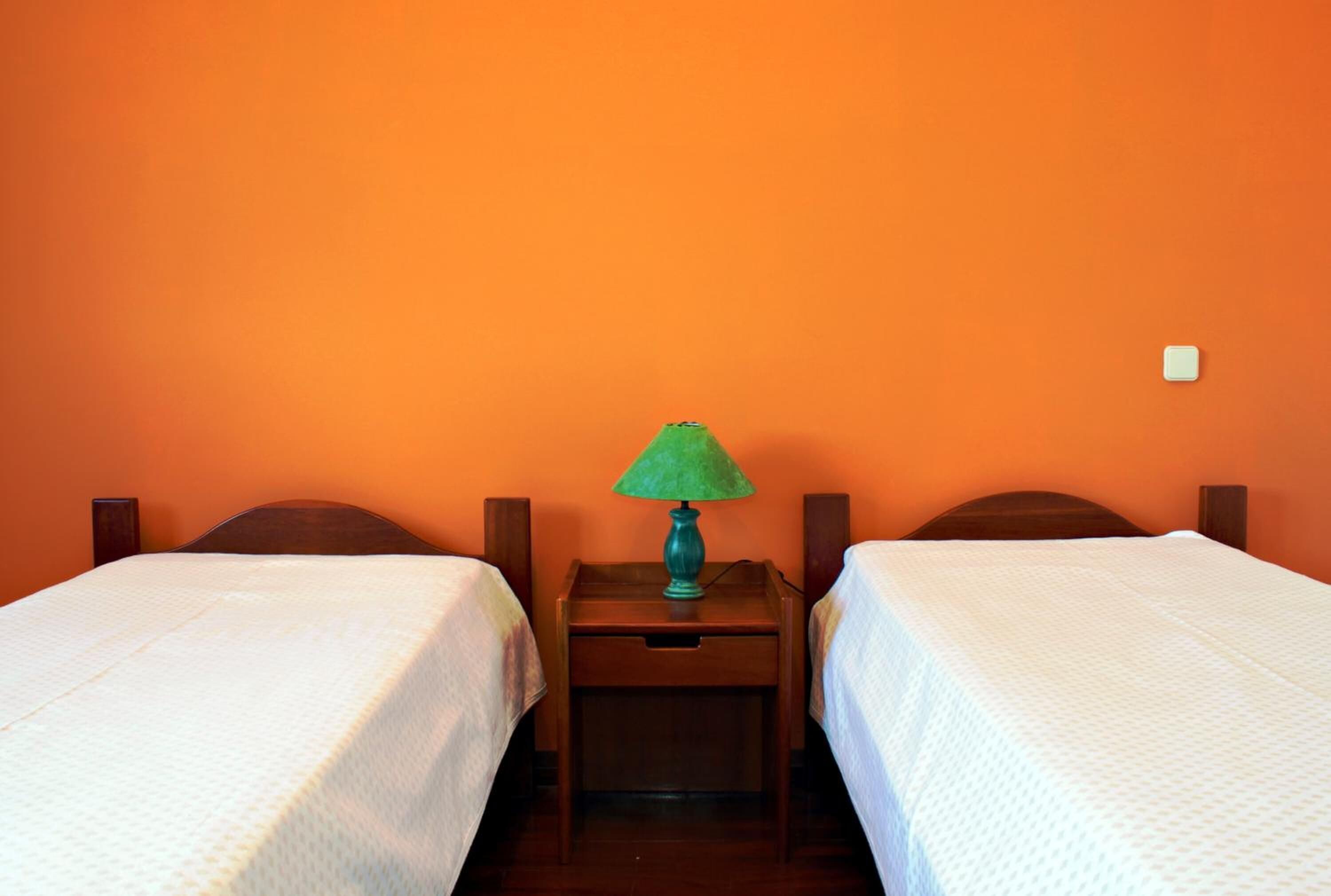 Quarto com duas camas de solteiro, mesa de cabeceira com luminária e parede laranja.