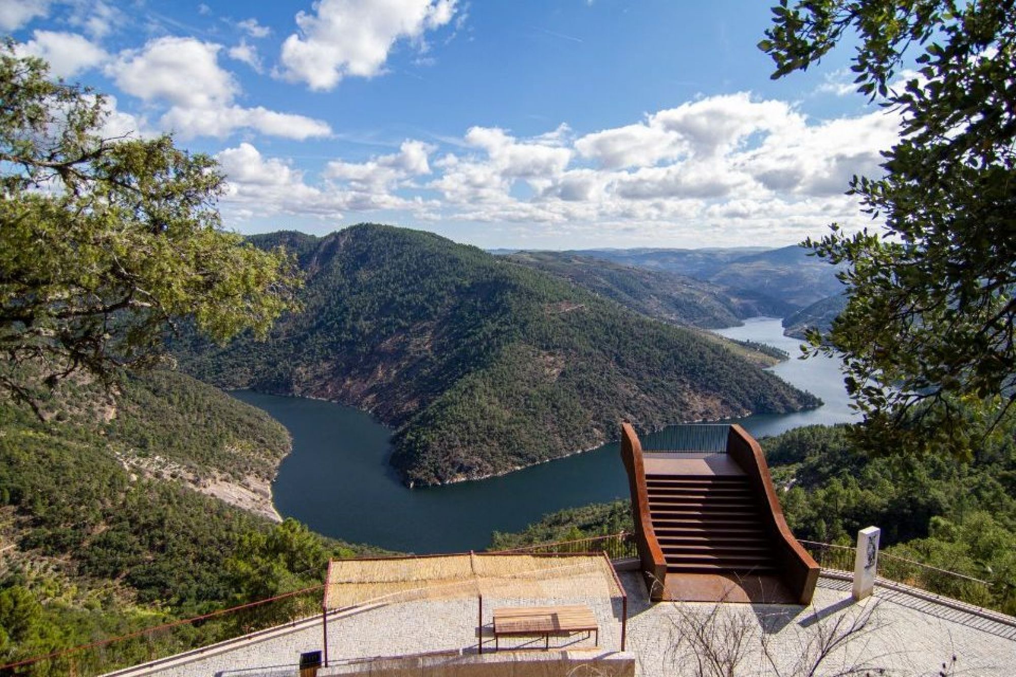 Um mirante panorâmico com uma escadaria que desce para uma plataforma de observação, com vista para um lago rodeado por colinas arborizadas.