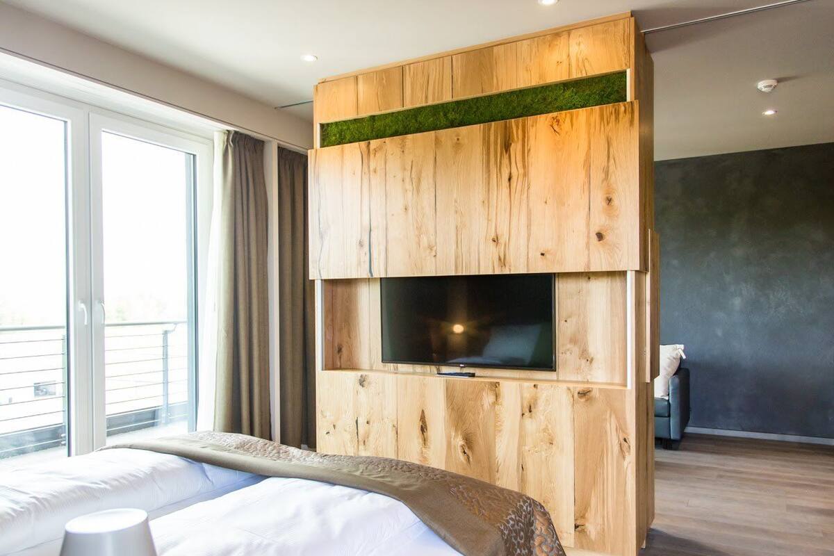 Quarto de hotel moderno com uma cama grande, televisão fixada numa parede revestida a madeira e uma varanda com cortinas.