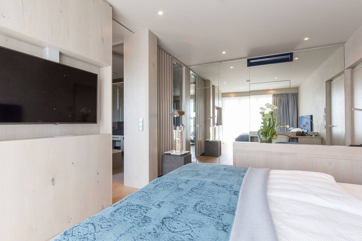 Quarto de hotel moderno com uma cama grande, uma TV de tela plana, uma escrivaninha com cadeira e um banheiro visível através de uma porta de vidro.
