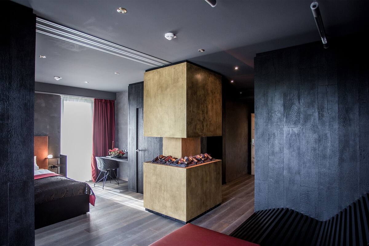 Quarto de hotel moderno com parede de destaque em madeira, lareira, cama e área de estar.