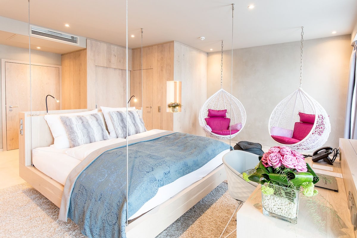 Um quarto moderno com cama, cadeiras suspensas, mesa de cabeceira com um vaso de flores e um telefone fixado na parede.