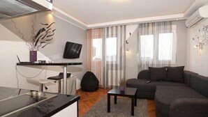 Uma sala de estar moderna com um sofá, uma mesinha e uma televisão. Há uma área de cozinha com bancada e uma cadeira.