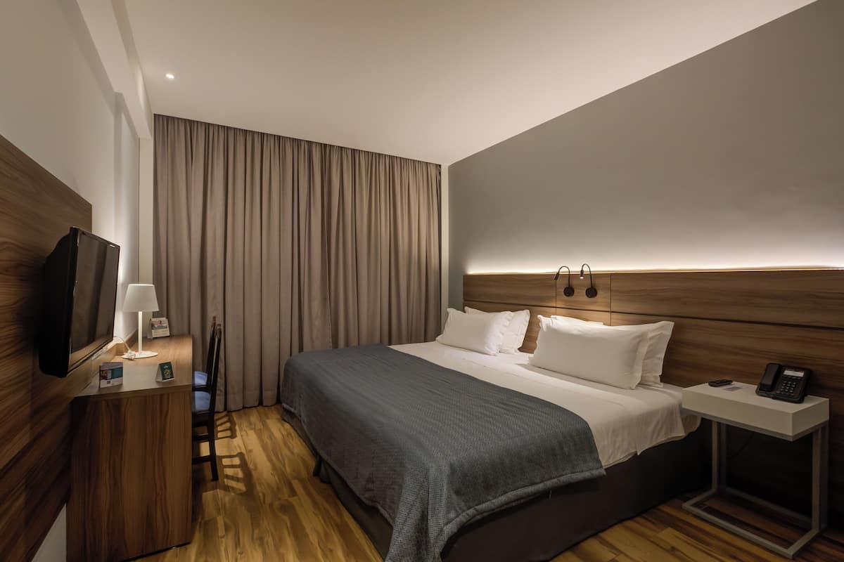 Quarto de hotel moderno com cabeceira de madeira, uma cama grande com lençóis brancos, mesa de cabeceira com luminária, televisão de tela plana e telefone.