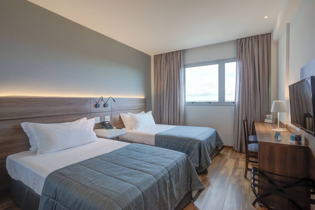 Quarto de hotel com duas camas, uma escrivaninha, televisão e janela com cortinas.