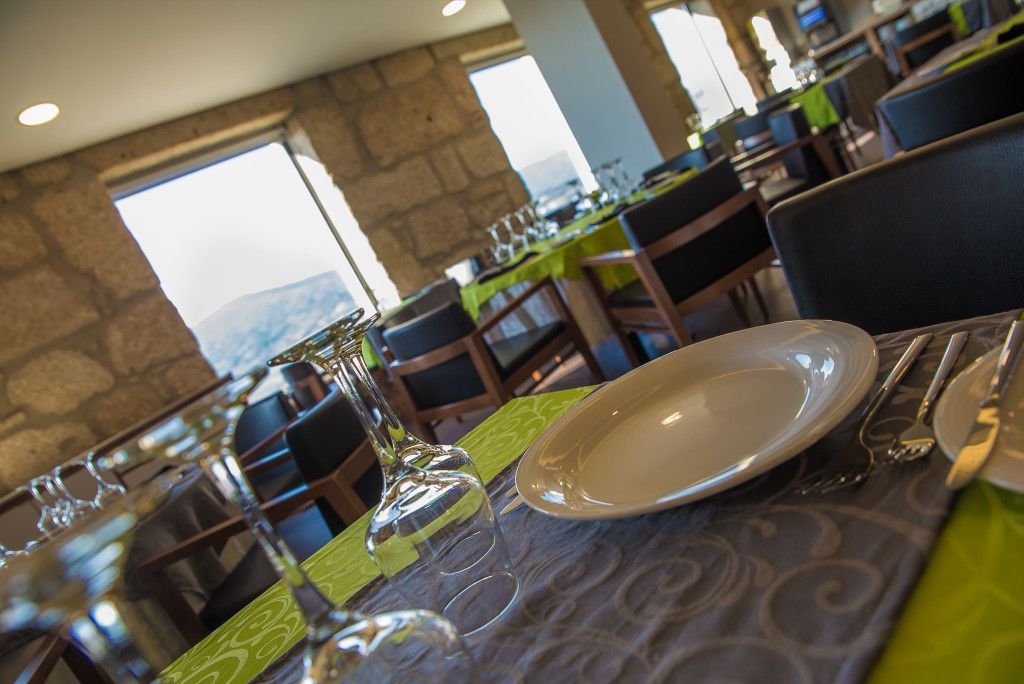 Ambiente de restaurante com mesas postas para refeições, incluindo copos e pratos transparentes, e vista para o exterior através de grandes janelas.