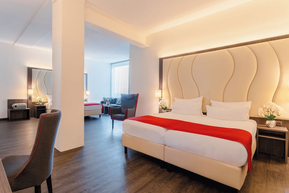 Quarto de hotel moderno com uma cama grande, uma escrivaninha e uma cadeira. Possui um espaço de estar separado com um sofá e poltronas.