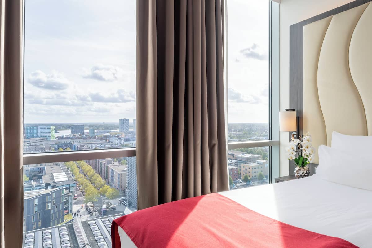 Quarto de hotel com uma cama grande, vista para a cidade e uma janela com cortinas.