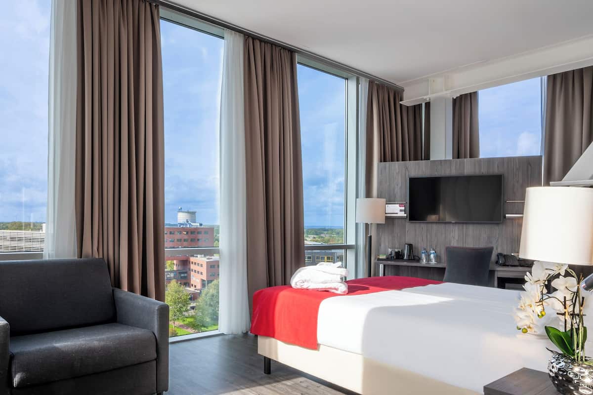 Quarto de hotel com uma cama grande, uma poltrona, uma escrivaninha, uma televisão e vista para a cidade.