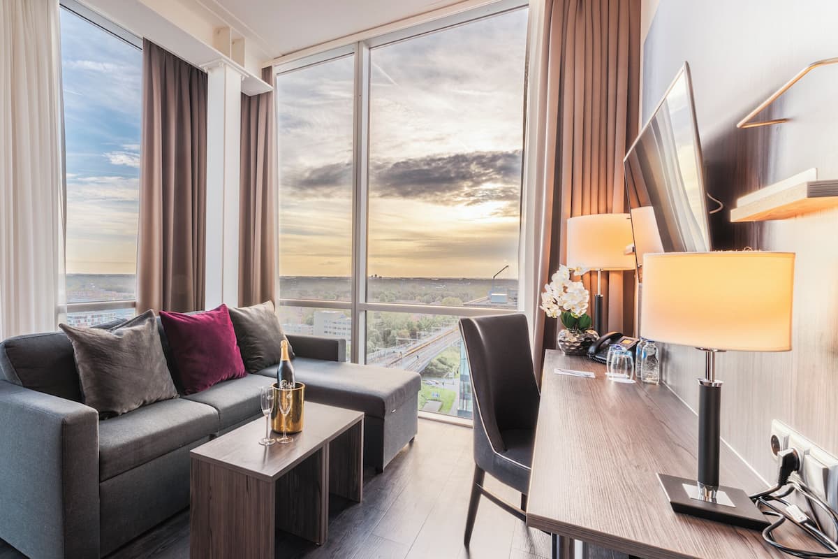 Quarto de hotel moderno com uma janela grande, um sofá, uma mesa pequena com garrafa e copo, uma cadeira e uma escrivaninha com abajur.