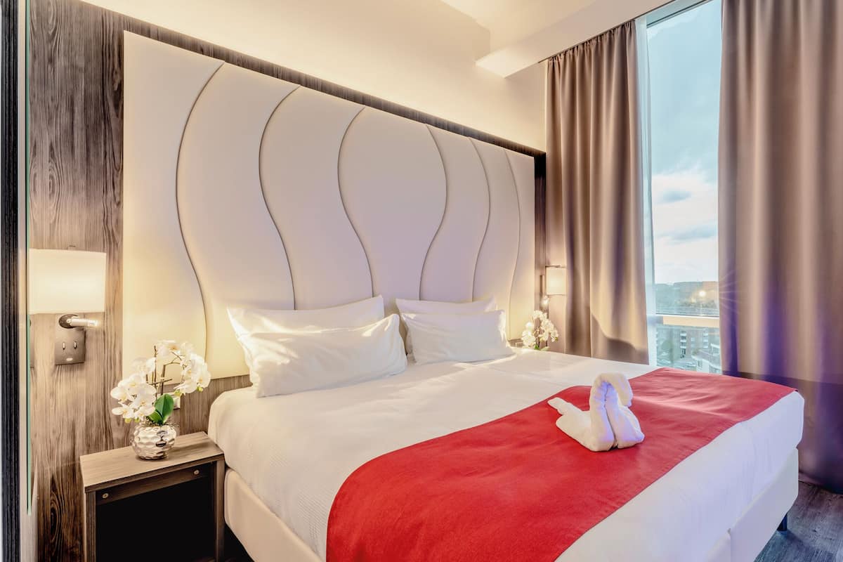 Quarto de hotel com uma cama grande, manta vermelha, almofadas brancas, cabeceira branca, mesa de cabeceira com um vaso de flores e vista para a cidade pela janela.