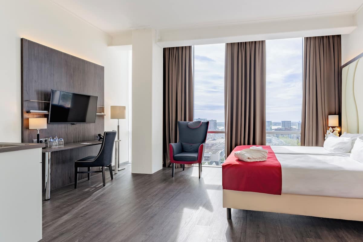 Quarto de hotel com cama, escrivaninha, cadeira, televisão e janelas amplas com cortinas.