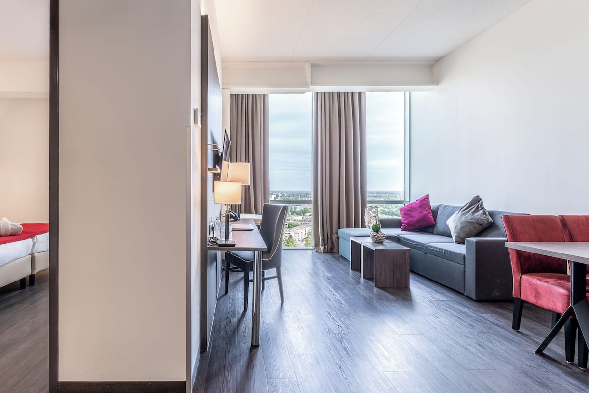 Quarto de hotel com cama, escrivaninha, sofá e mesa de jantar. Há uma janela grande com vista para a cidade.