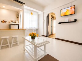 Cozinha moderna com armários brancos, forno embutido e uma geladeira pequena. Há uma área de refeições com uma mesa de vidro e dois banquinhos brancos. Um vaso de flores está sobre a mesa, e uma TV de tela plana está fixada na parede.