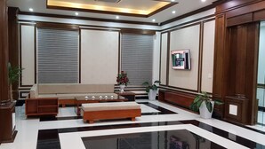 Quarto de hotel moderno com um sofá, uma mesa de centro e uma mesinha lateral. Há duas plantas em vasos e uma TV fixada na parede.