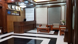 Uma área de recepção de hotel com um balcão de madeira, vários relógios e um espaço de estar com sofás e uma mesa de centro.