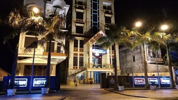 Um hotel de vários andares com entrada central, palmeiras e placas iluminadas com os dizeres 'HOTEL' e 'CAFÉ'.