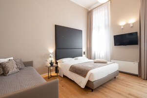 Quarto de hotel moderno com uma cama grande, um sofá menor, uma mesa de cabeceira com abajur, uma TV de tela plana na parede e uma janela grande com cortinas.