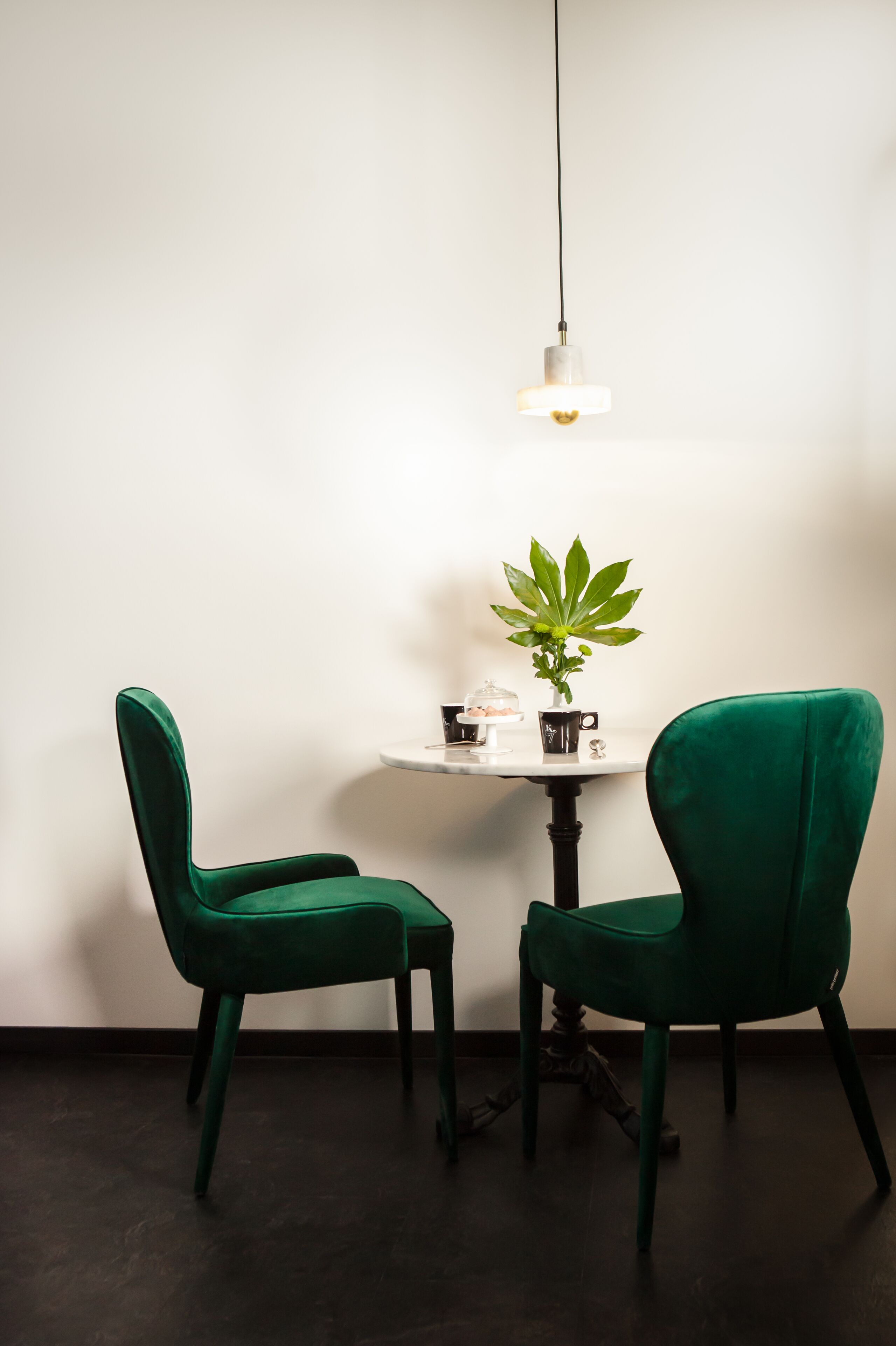 Uma mesa pequena e redonda com duas cadeiras verdes, um vaso de planta e um candeeiro suspenso.