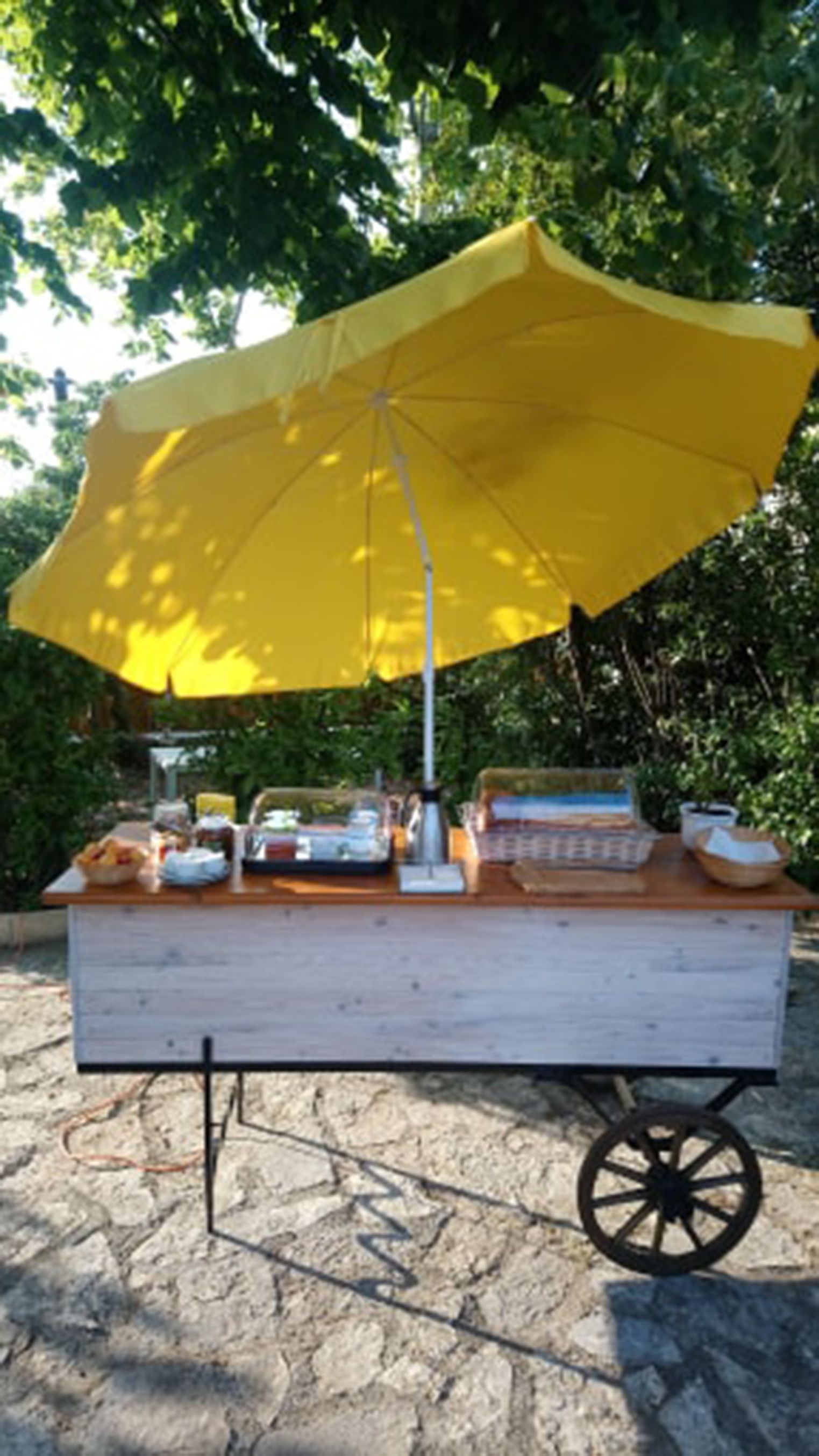 Um carrinho de madeira com guarda-sol amarelo, alimentos e um caixa registradora.