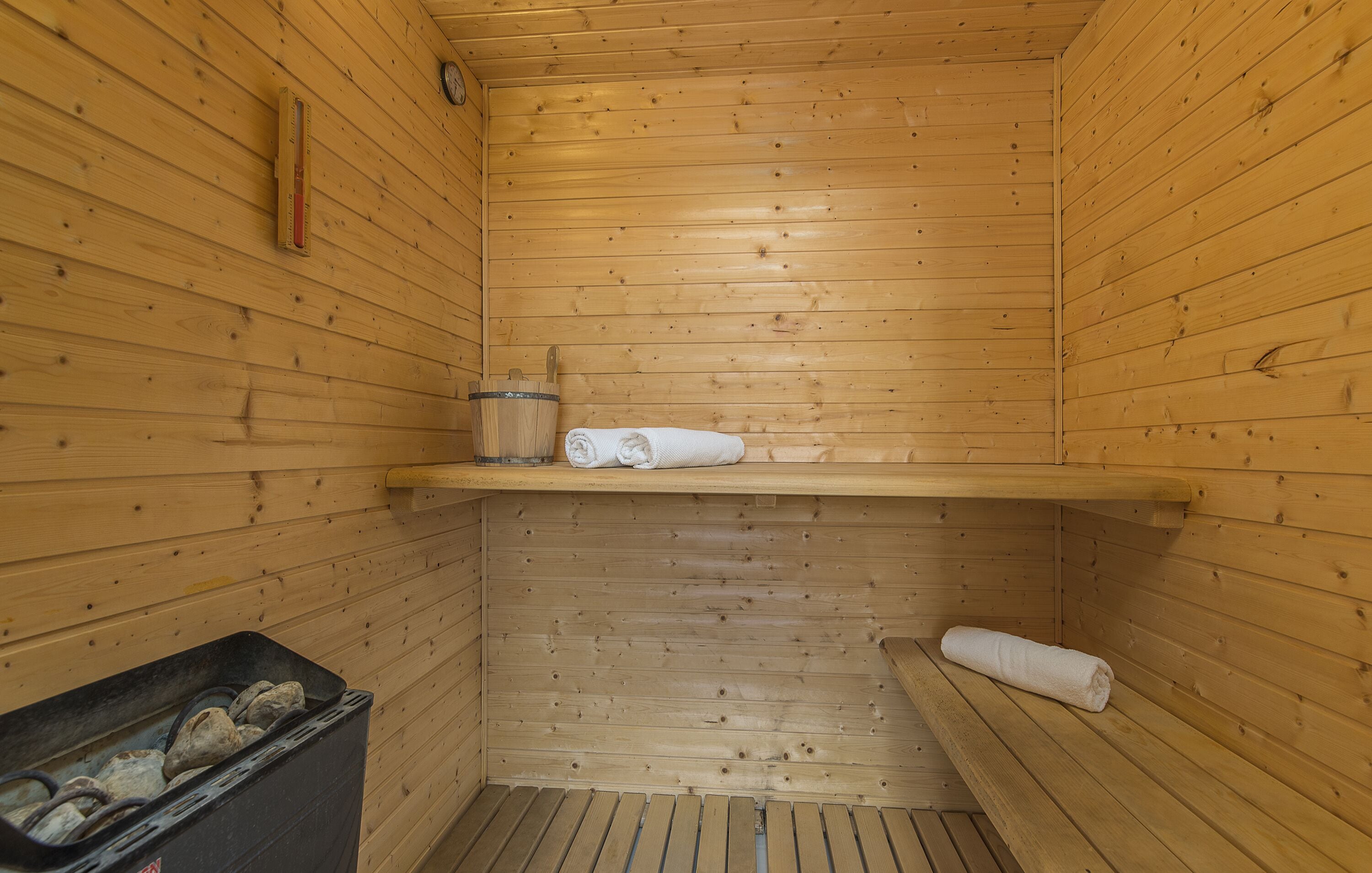 Uma sala de sauna com paredes de madeira, um banco, uma prateleira com toalhas e um recipiente com pedras.
