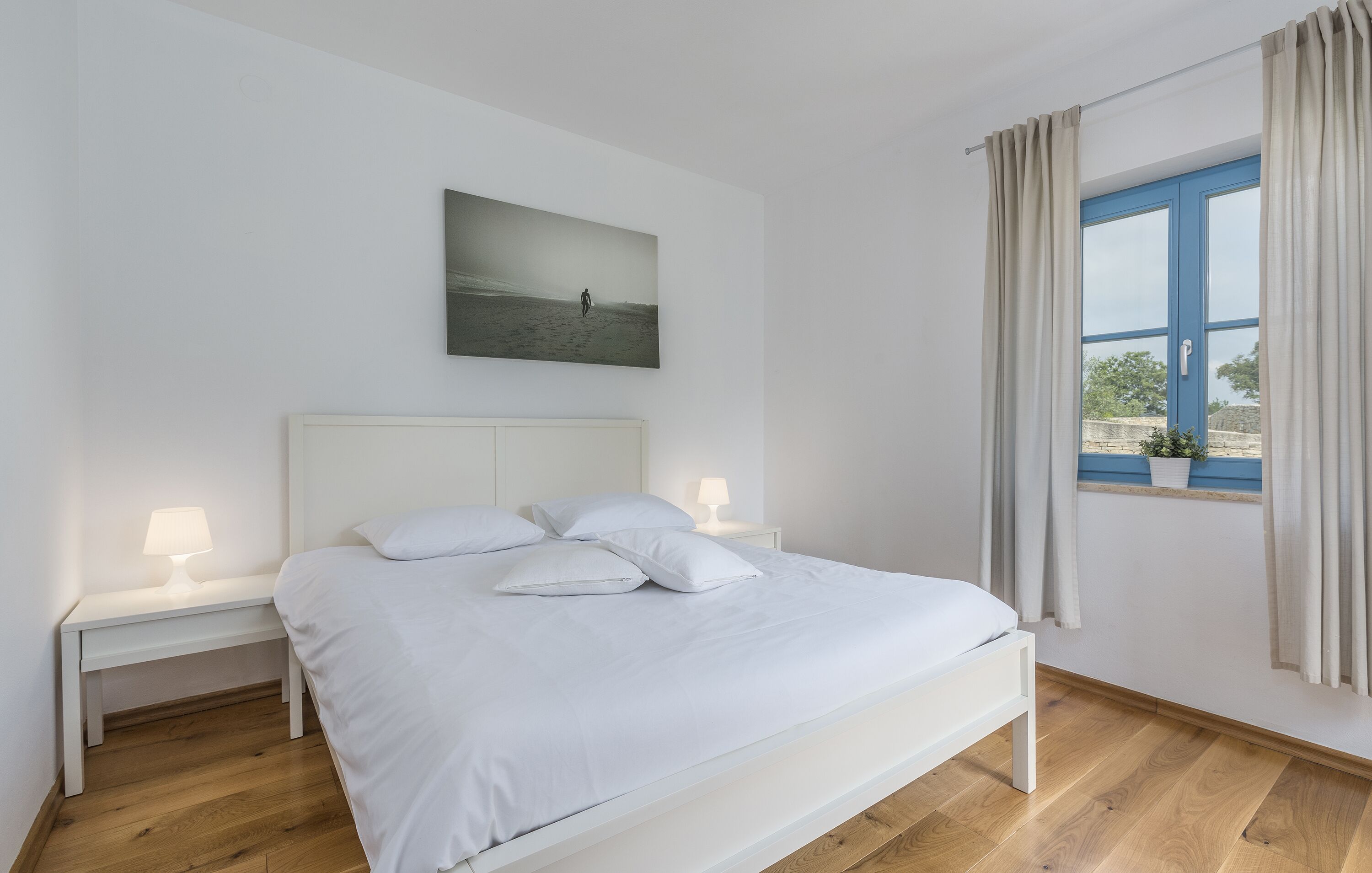 Quarto com cama, mesas de cabeceira, um criado-mudo, janela com cortinas e um quadro na parede.