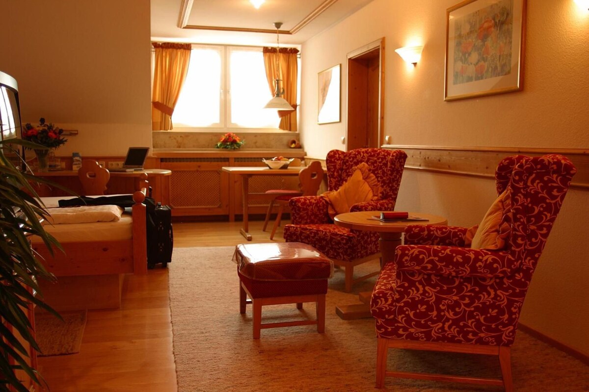 Uma sala de estar acolhedora com sofá, poltrona e apoio para os pés, uma área de refeições com mesa e cadeiras, e uma cozinha ao fundo.