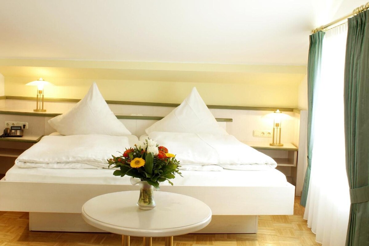 Quarto de hotel com uma cama grande, duas mesas de cabeceira, uma mesinha redonda com um vaso de flores e piso de madeira.